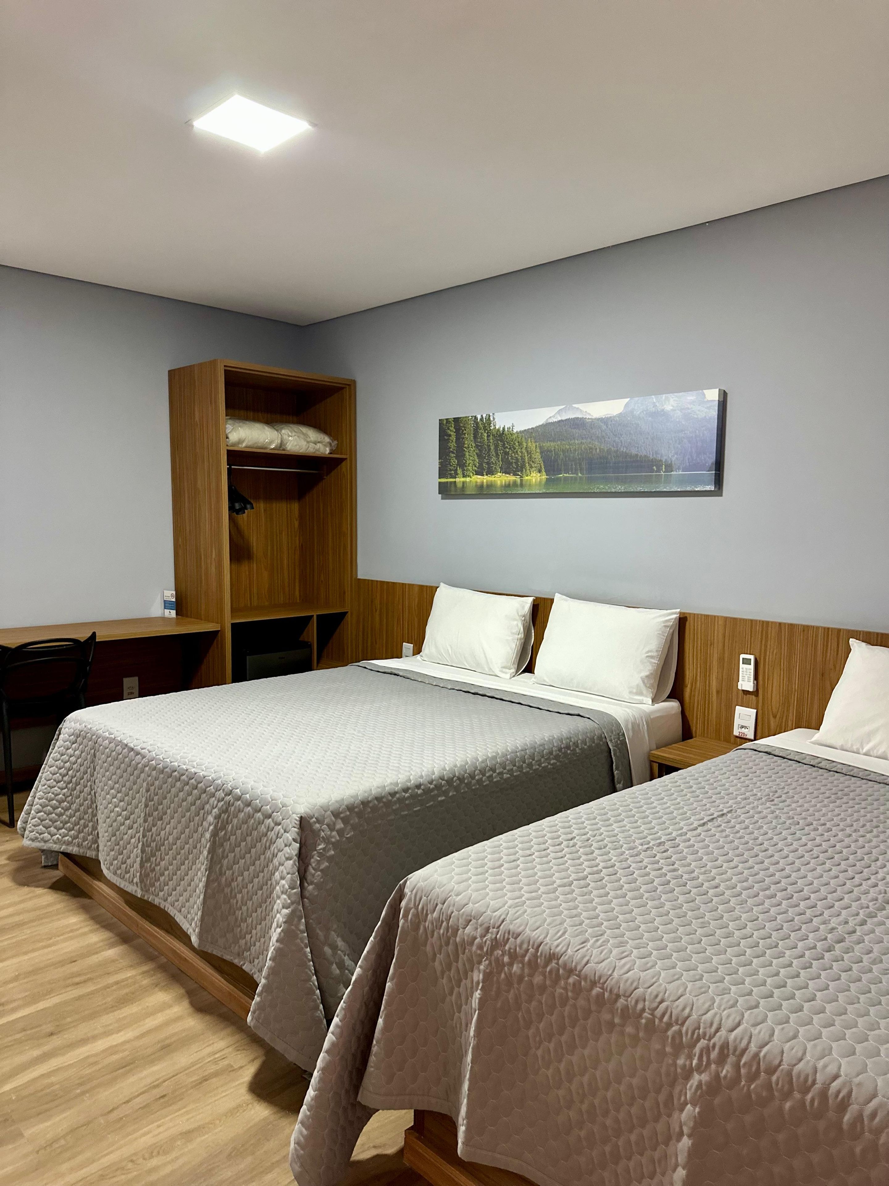 Quarto de hotel com duas camas, guarda-roupa de madeira, escrivaninha e um quadro na parede.