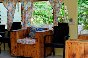 Área de estar ao ar livre com bancos de madeira, uma cadeira e uma mesa, cercada por uma parede amarela e plantas verdes.