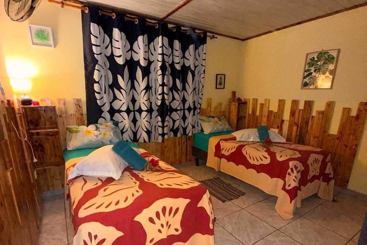 Quarto com duas camas, cabeceira de madeira, ventoinha, cortina estampada com folhas e uma pequena imagem na parede.