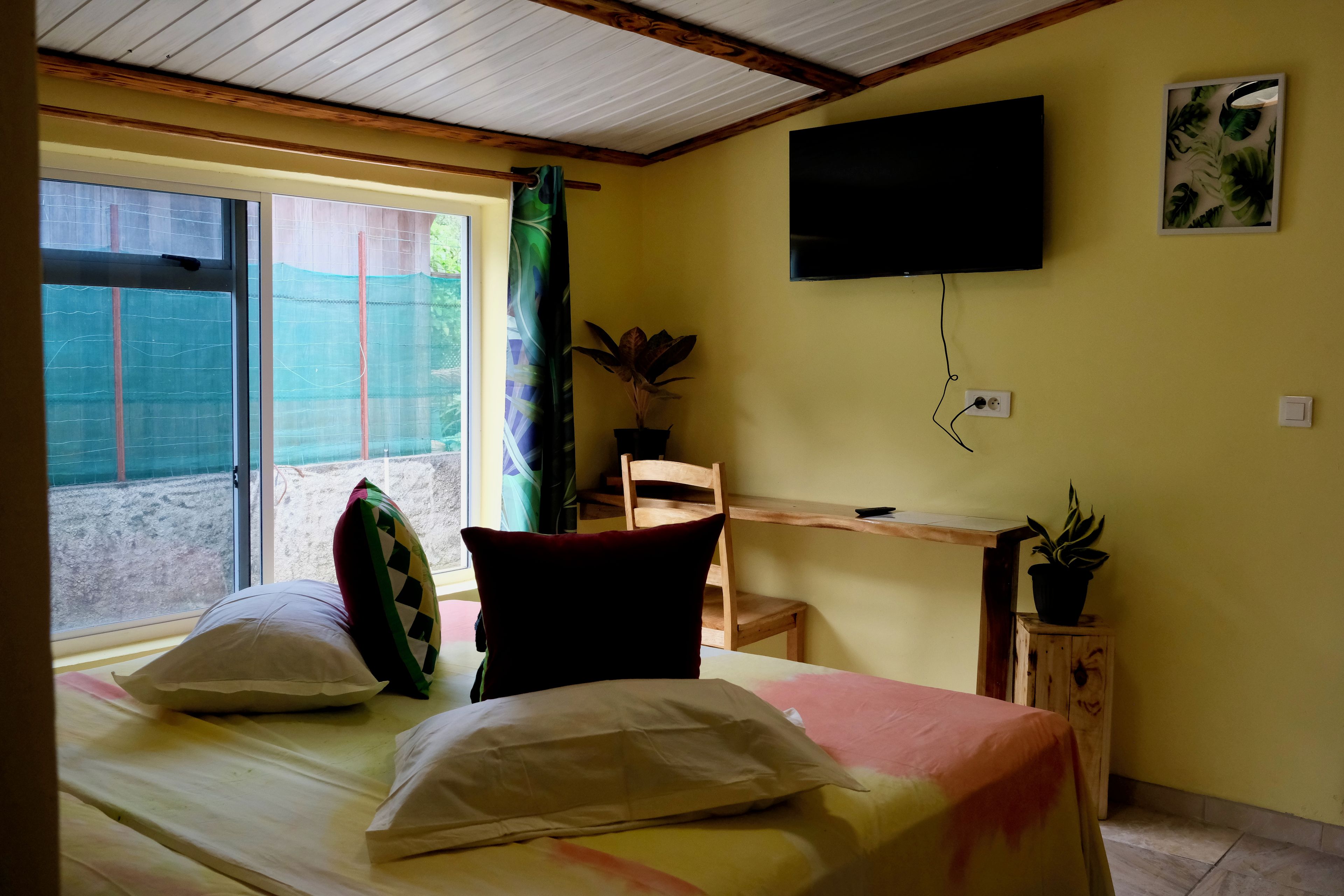 Quarto com cama, escrivaninha, cadeira, televisão e janela com vista para o exterior.
