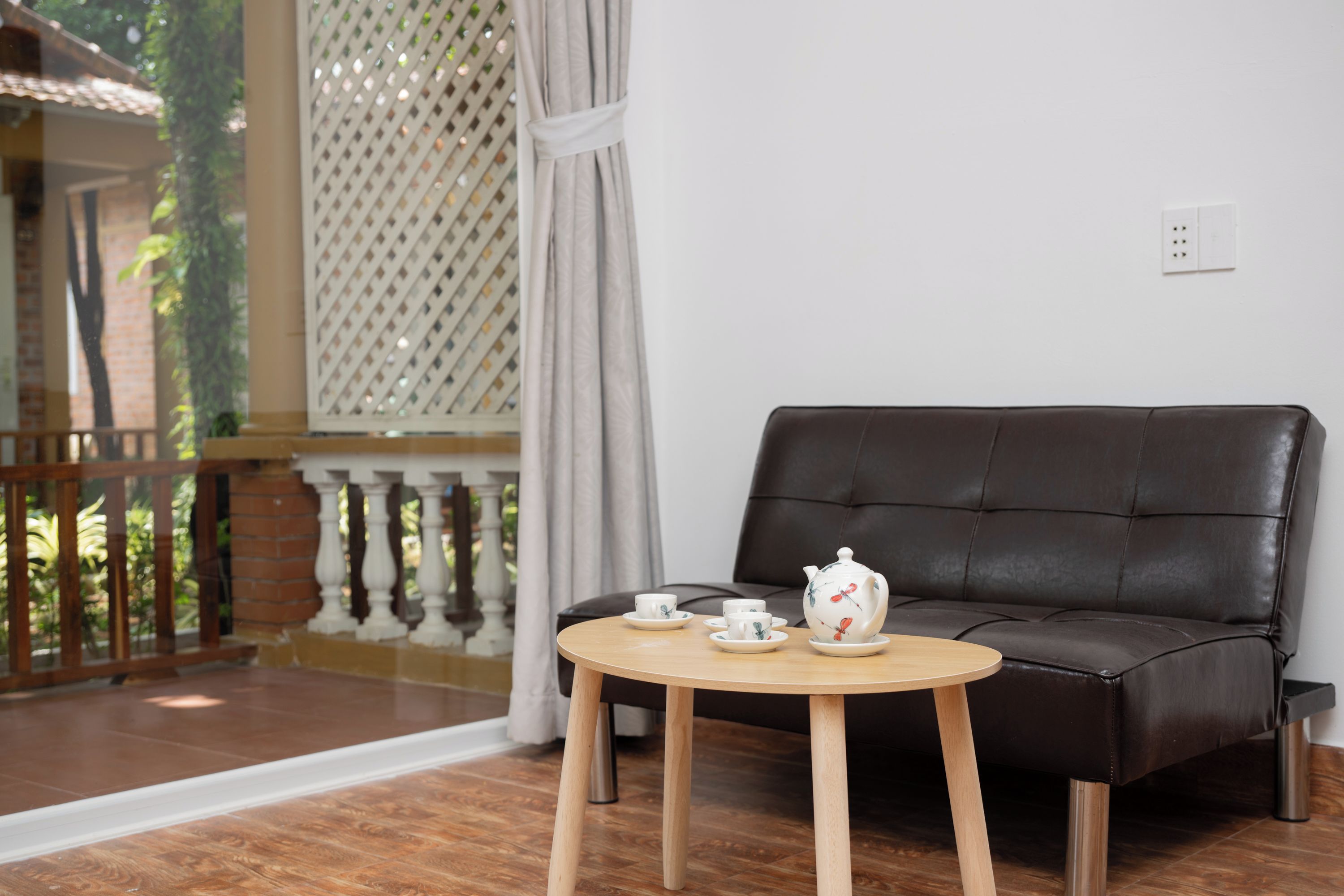 Quarto com sofá de couro marrom, mesa de madeira com bule e xícaras, e vista para área verde lá fora.