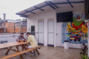 Uma área de estar externa com mesa e bancos de madeira, um mural com arte colorida na parede e uma televisão fixada na parede.