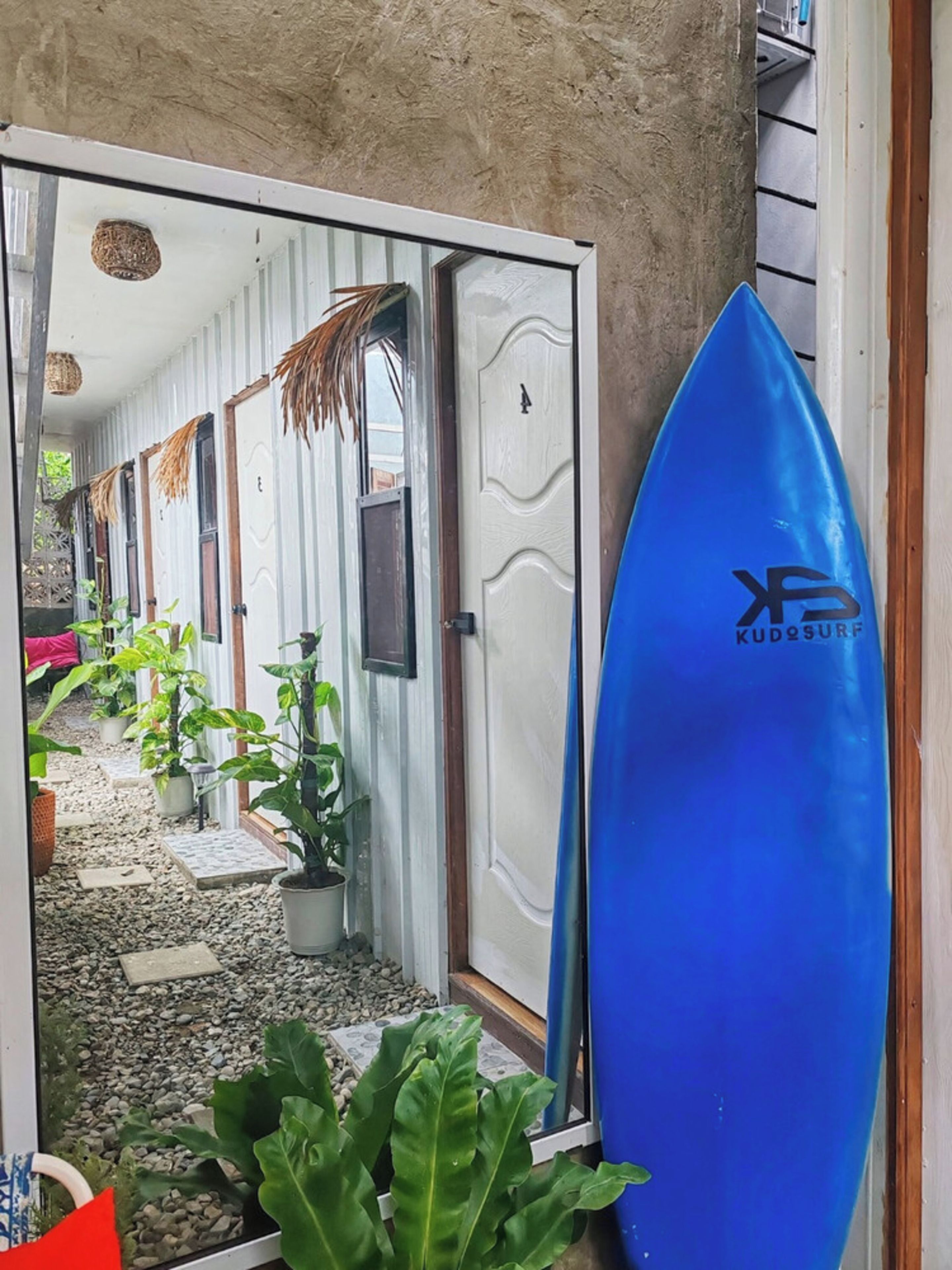 Corredor com portas, plantas em vasos e uma prancha de surf azul encostada na parede.