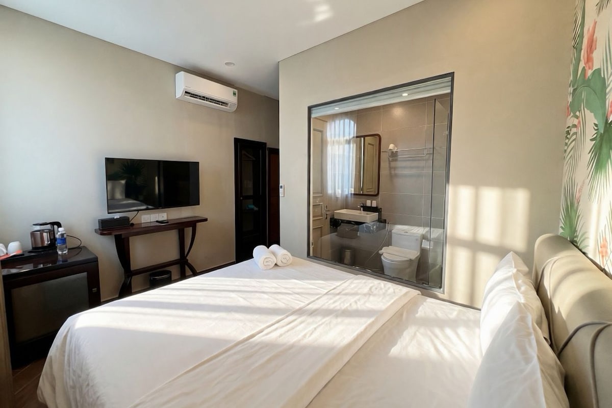 Quarto de hotel com cama, escrivaninha com televisão, espelho e banheiro visível através de um painel de vidro.