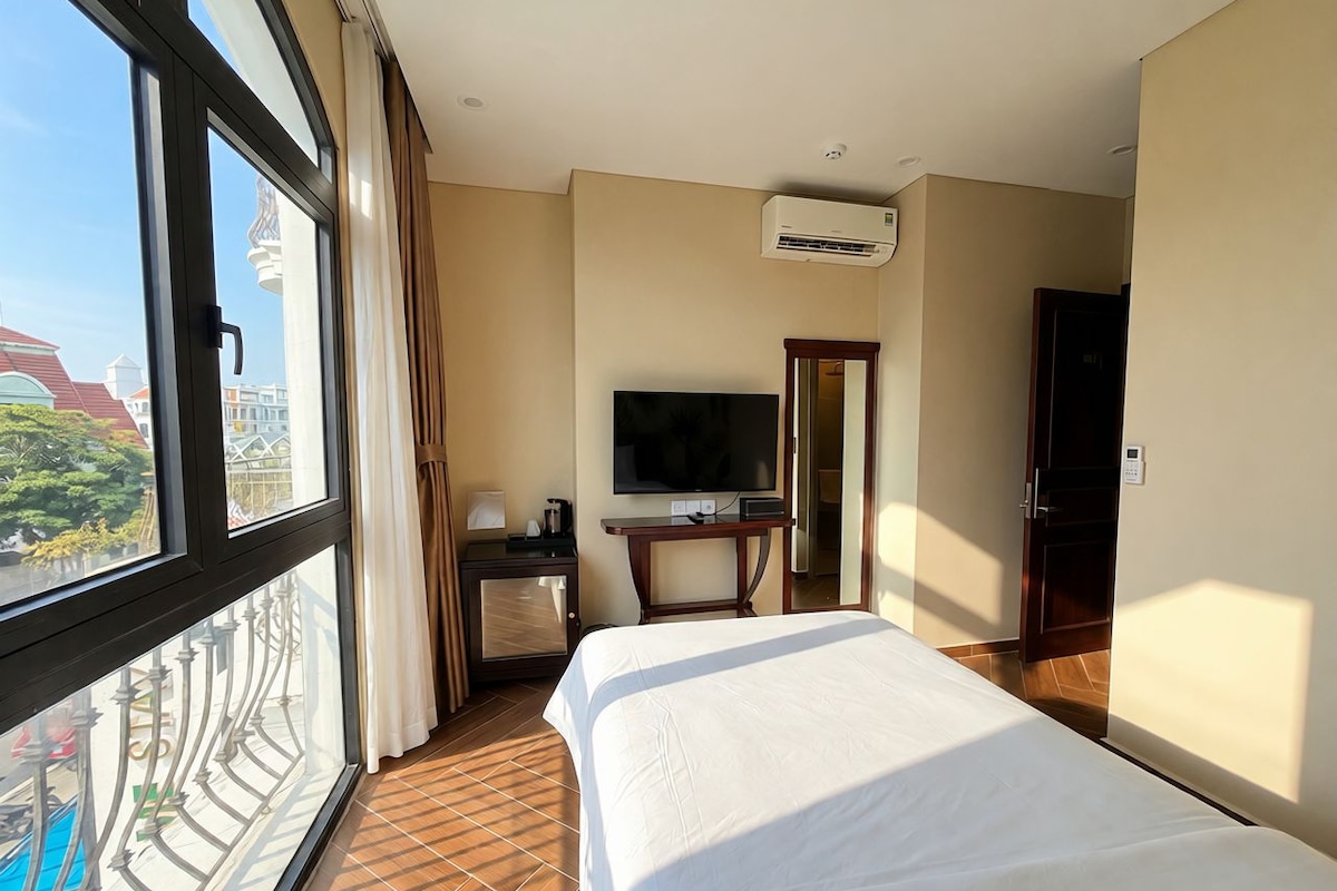 Quarto de hotel com uma cama grande, televisão, escrivaninha e vista da cidade através de amplas janelas.