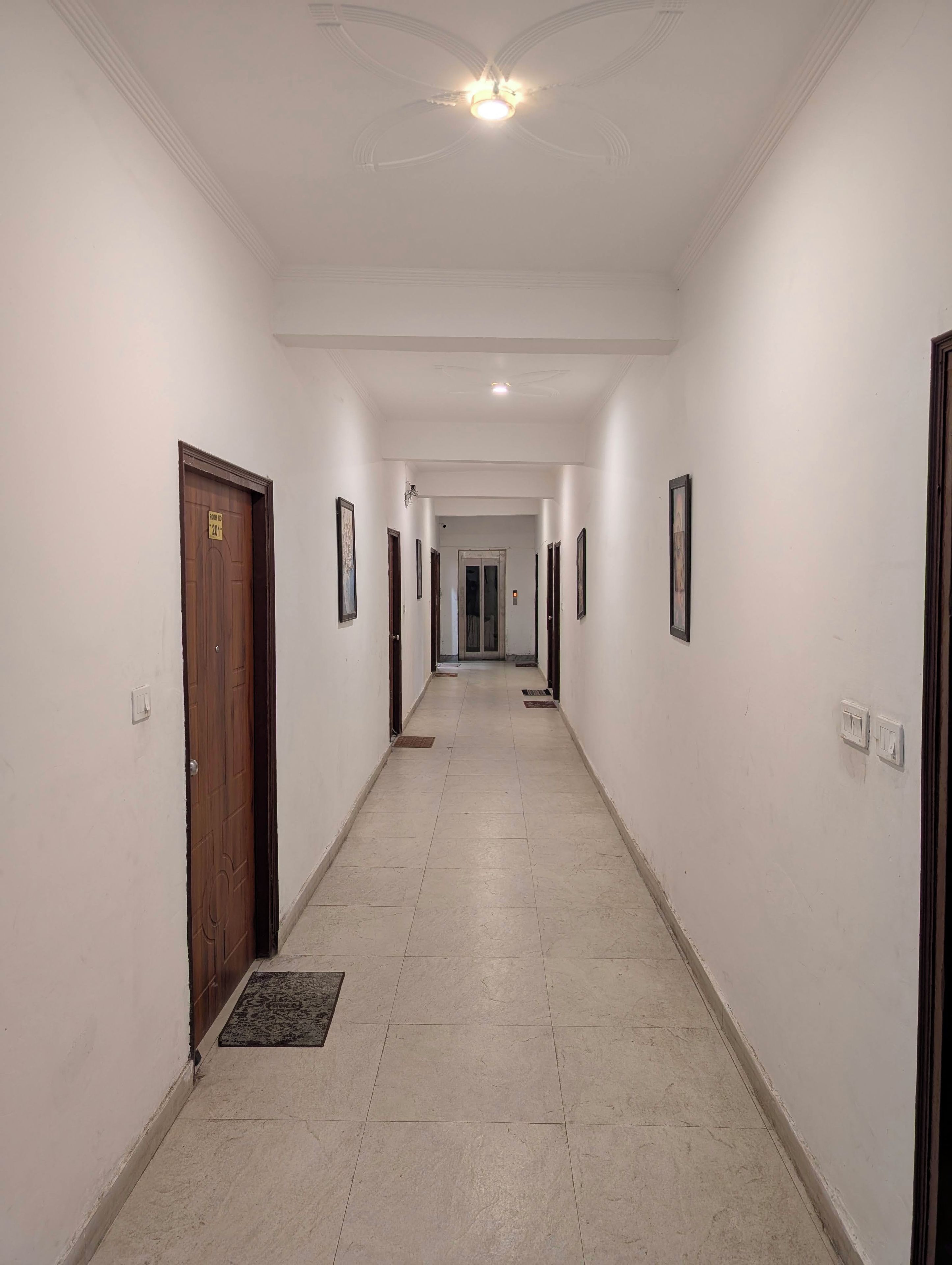 Um corredor comprido e estreito com paredes brancas, uma porta de madeira à esquerda e um ventilador de teto no final.