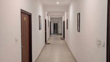 Um corredor comprido e estreito com paredes brancas, uma porta de madeira à esquerda e um ventilador de teto no final.
