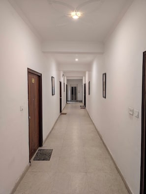 Um corredor comprido e estreito com paredes brancas, uma porta de madeira à esquerda e um ventilador de teto no final.