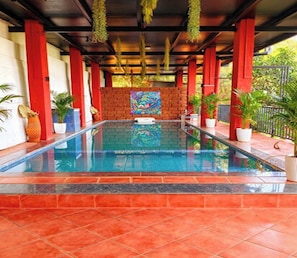 Piscina com piso ladrilhado, rodeada por pilares vermelhos e uma parede de tijolos com um mural colorido.