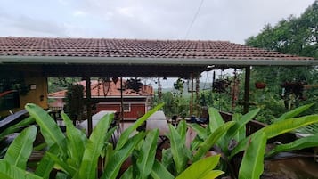 Área externa coberta com telhado de azulejos, cercada por plantas verdes exuberantes.