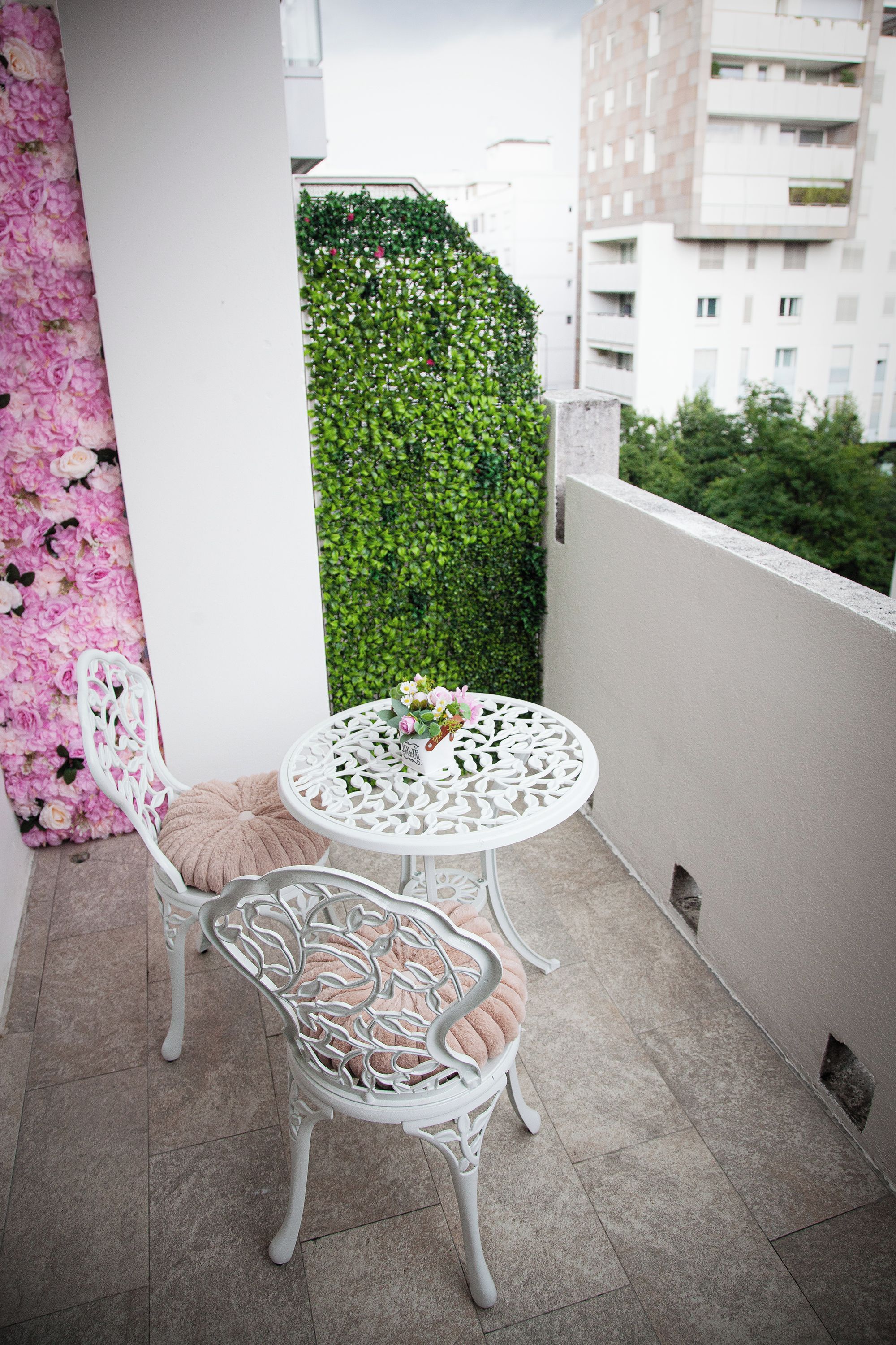 Uma varanda com mesa e cadeiras brancas, um muro com flores cor-de-rosa e uma sebe verde.