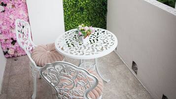 Uma varanda com mesa e cadeiras brancas, um muro com flores cor-de-rosa e uma sebe verde.
