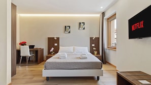 Quarto de hotel moderno com cama, escrivaninha, poltrona, televisão e obras de arte na parede.