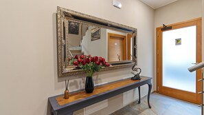 Um corredor com um espelho grande e ornamentado, uma mesa consola de madeira com um vaso de rosas vermelhas e uma porta com uma placa.