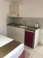 Quarto de hotel compacto, com uma cama, cozinha compacta com pia e chaleira, e um pequeno armário sobre a pia.