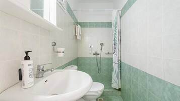 Casa de banho com lavatório branco, sanita e chuveiro com parede revestida a mosaico verde e branco.