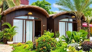 Duas casas de campo com telhados de palha e grandes janelas, rodeadas por vegetação exuberante e plantas tropicais.