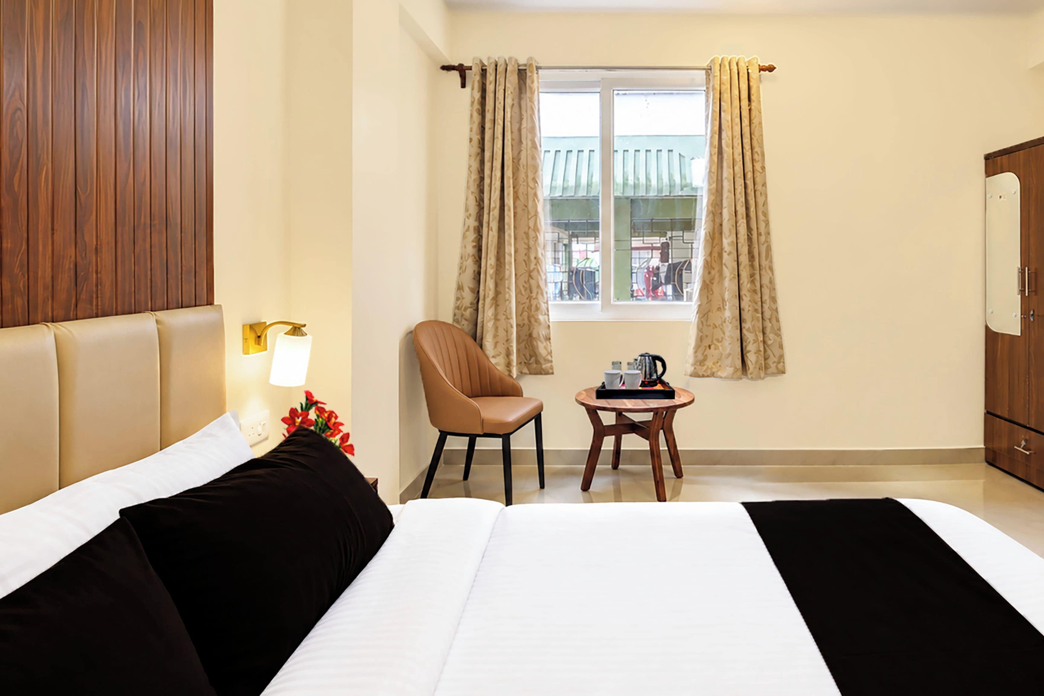 Quarto de hotel com cama, uma cadeira, uma mesinha com bandeja, janela com cortinas e uma luminária na parede.