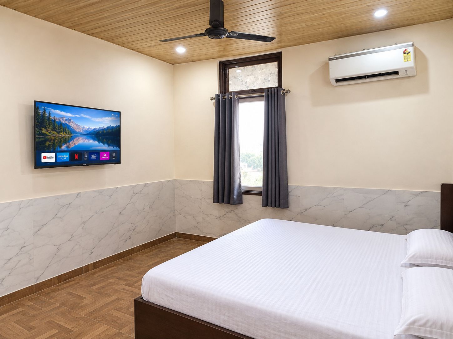 Quarto de hotel com cama, ventoinha no teto, aparelho de ar condicionado, televisão fixada na parede e janela com cortinas.