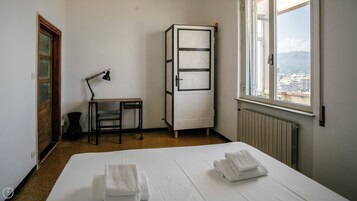 Quarto de hotel com uma cama, uma escrivaninha com cadeira, um radiador e uma janela com vista para edifícios e montanhas.