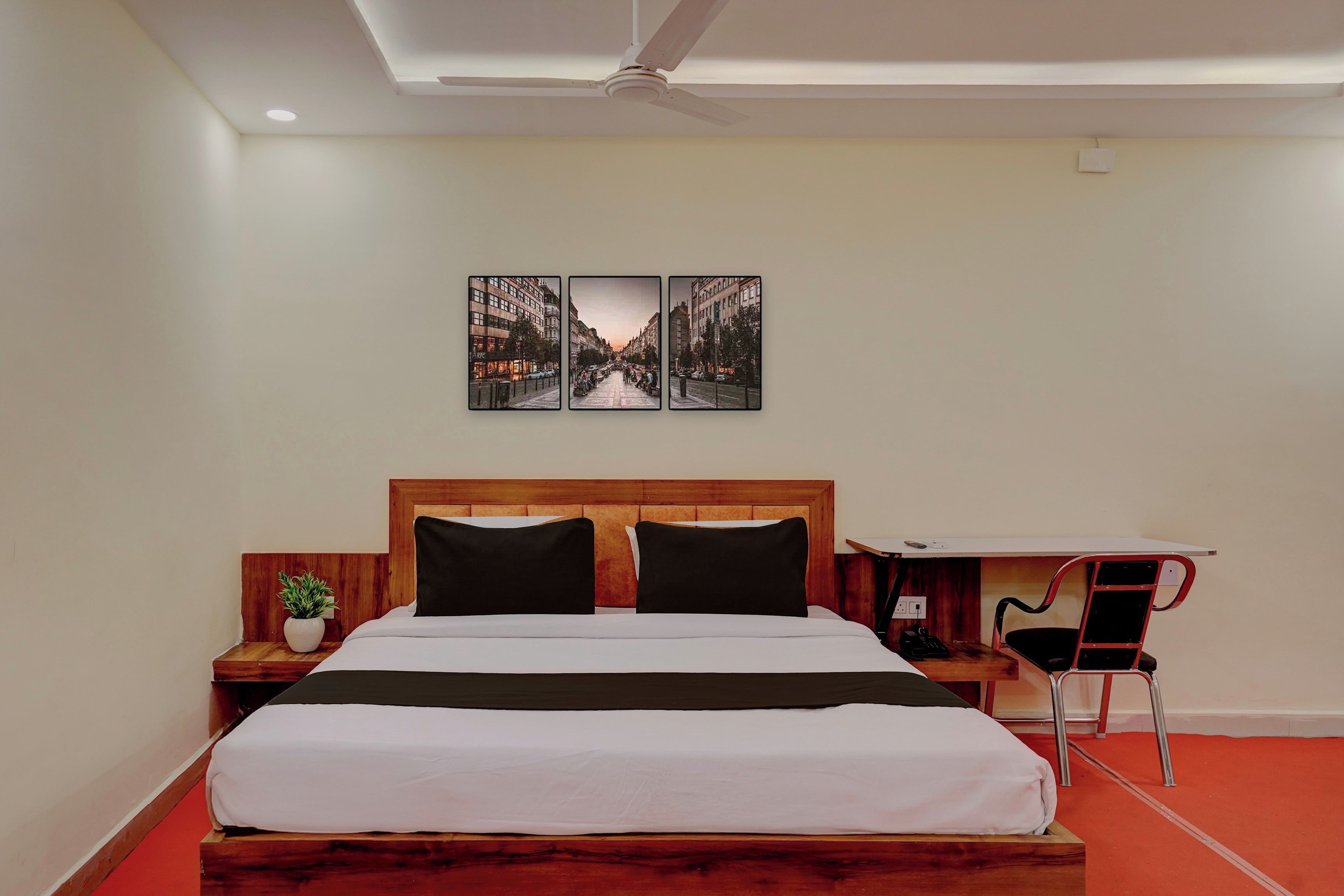 Quarto de hotel com cama, escrivaninha, cadeira, planta e obra de arte na parede.