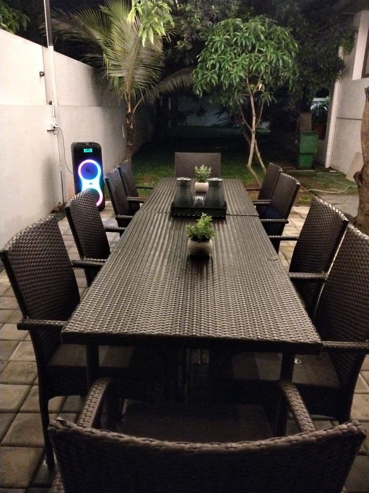 Área de refeições ao ar livre com uma mesa comprida e cadeiras, rodeada por vegetação e uma parede branca.