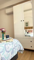 Quarto com cama, mesa de cabeceira com livros e um vaso de flores, guarda-roupa e espelho refletindo uma cama e um quadro.
