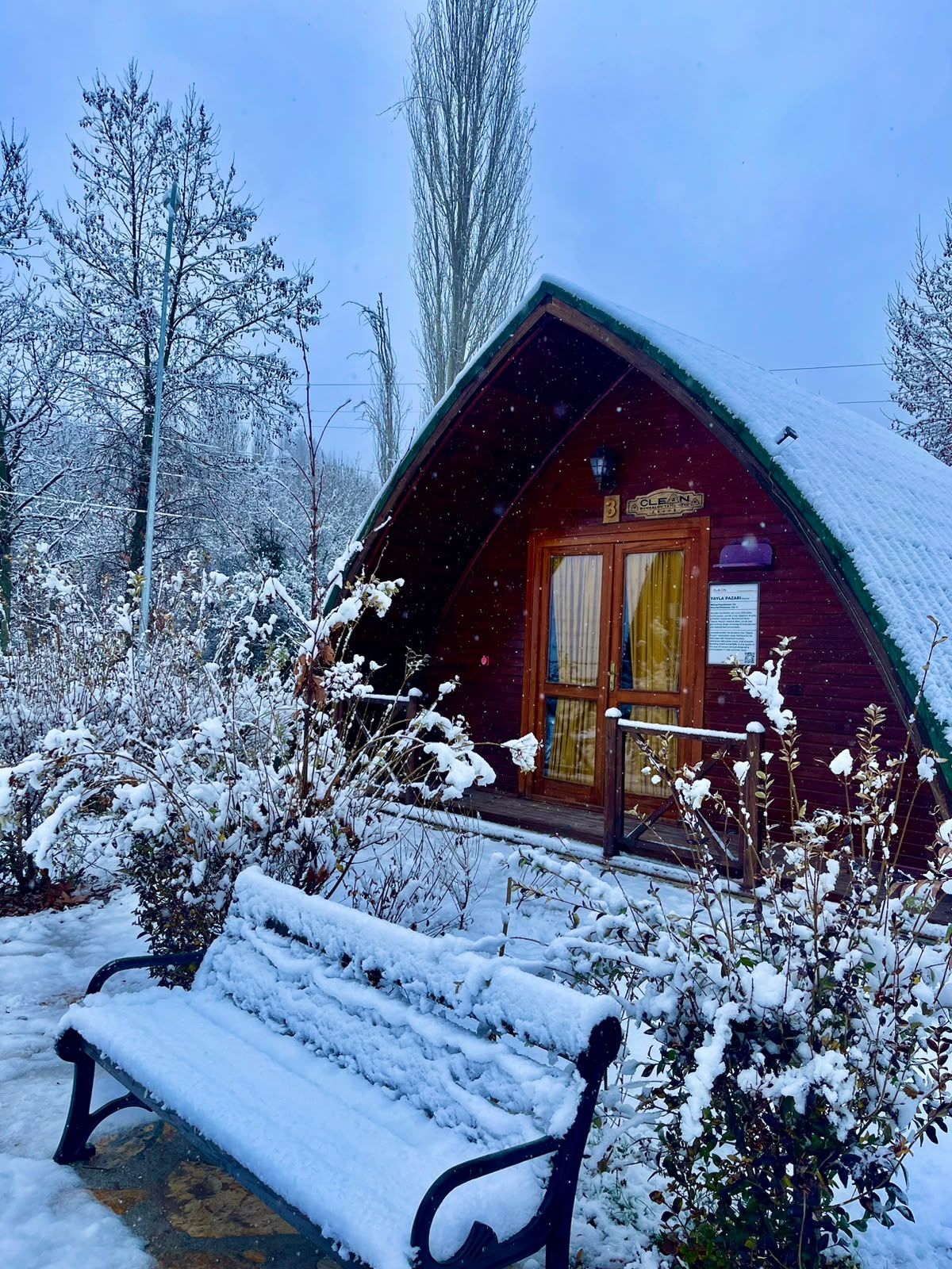 Uma cabana de madeira com telhado coberto de neve e um banco em frente.