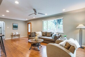 Uma sala de estar ampla com piso de madeira, um ventilador de teto, uma janela grande e uma área de estar com sofá e poltronas.