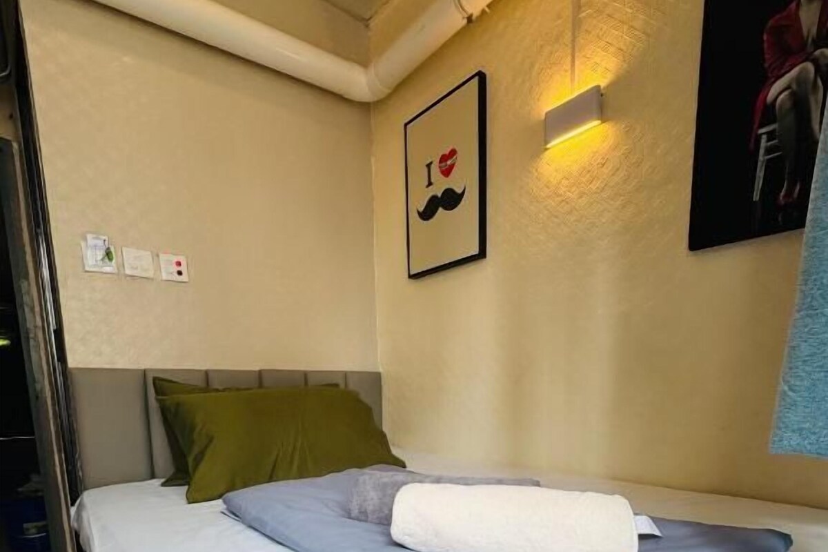 Uma cama de solteiro com colchão cinza, cobertor branco e travesseiro verde. Há um quadro na parede com um coração e um bigode.