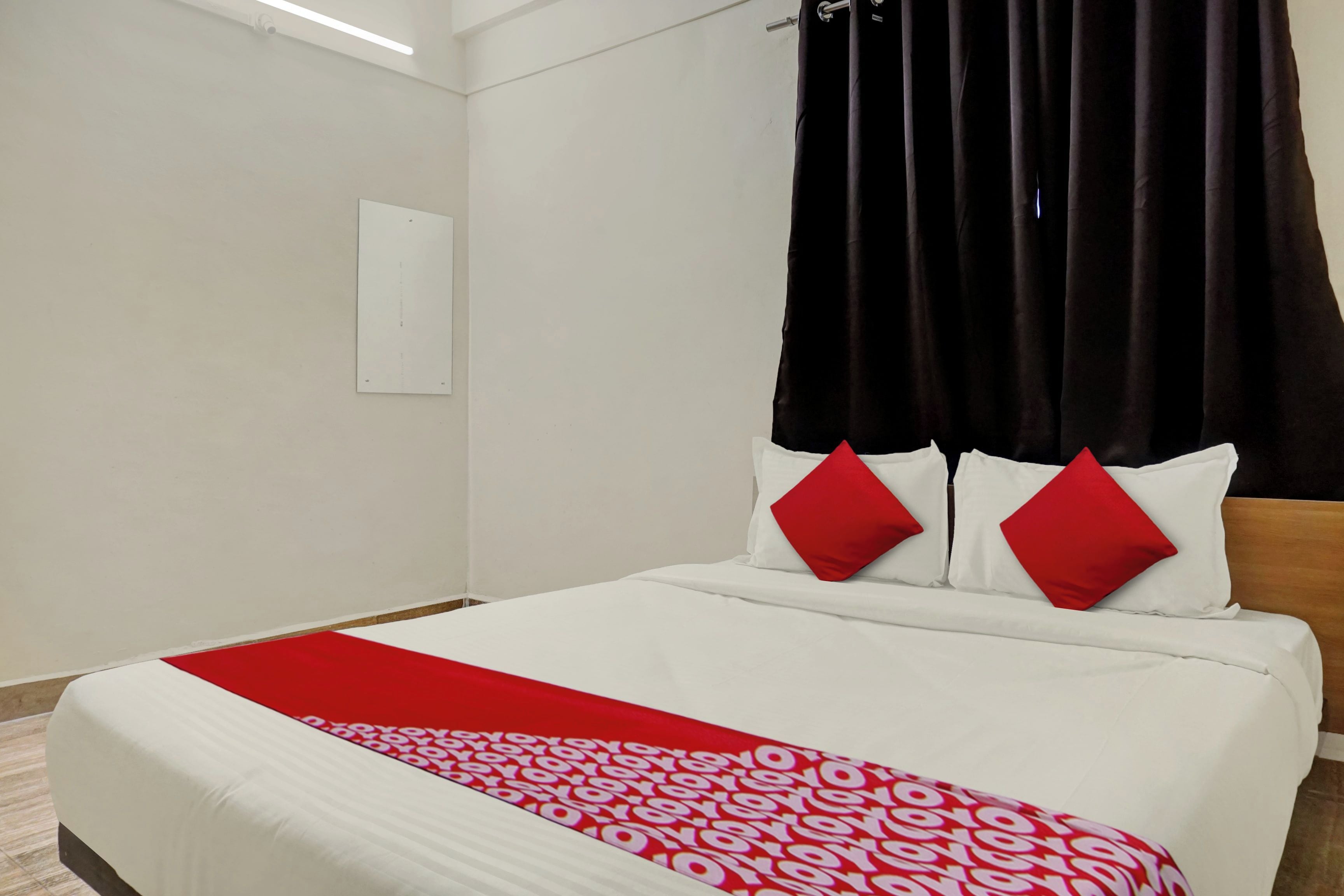Quarto de hotel com cama, colcha vermelha e branca estampada e cortinas pretas.