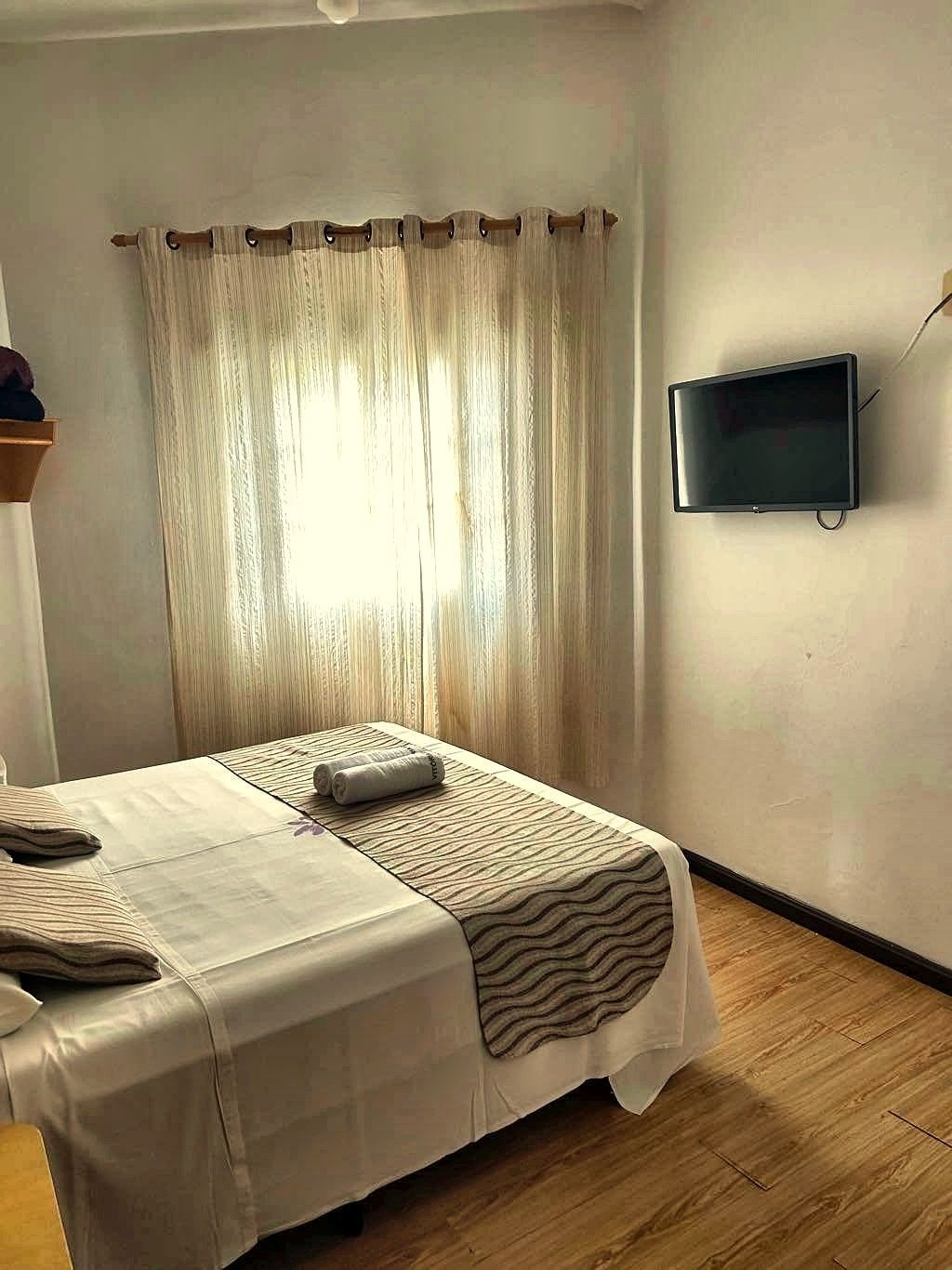 Quarto de hotel com uma cama, televisão presa à parede e uma janela com cortinas.