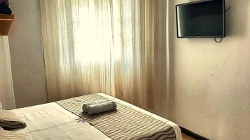 Quarto de hotel com uma cama, televisão presa à parede e uma janela com cortinas.