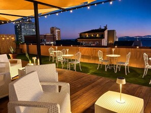 Área de estar externa com móveis de vime e mesas, com vista para o horizonte urbano ao entardecer.