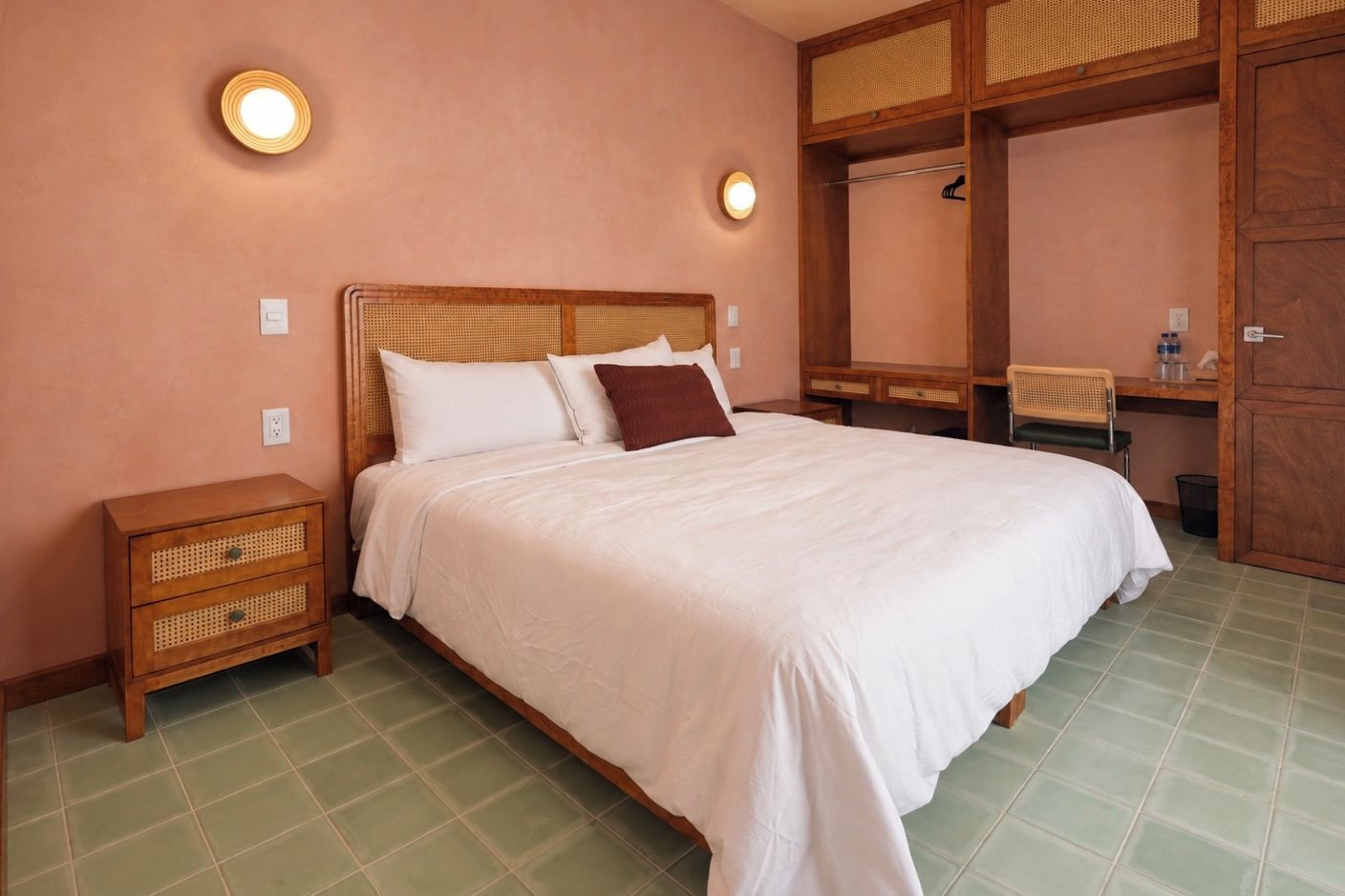 Uma cama bem arrumada com cabeceira, mesa de cabeceira com gavetas, uma cadeira e um guarda-roupa com espelho.