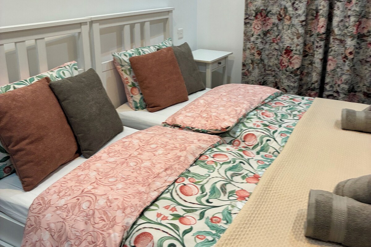 Quarto com duas camas, cada uma com colcha florida e várias almofadas. Há uma pequena mesa de cabeceira com abajur na parede.
