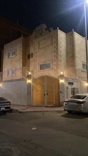 Um edifício de vários andares com entrada central, portão na entrada e dois carros estacionados.