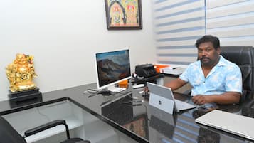 Um homem sentado em uma mesa com um laptop, um monitor de computador e um celular. Há uma estátua dourada na mesa e um quadro emoldurado na parede.