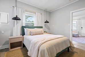 Quarto com cama, mesas de cabeceira, janela com cortinas e porta de acesso a outro cômodo.