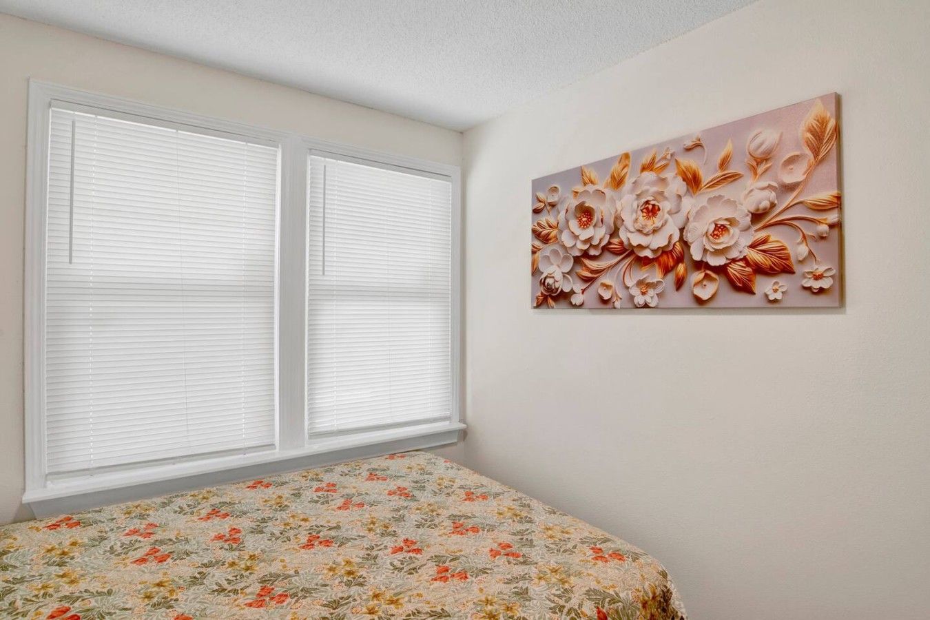 Quarto com colcha florida, uma janela grande com persianas e uma obra de arte floral na parede.