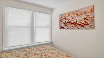 Quarto com colcha florida, uma janela grande com persianas e uma obra de arte floral na parede.