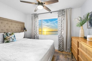 Quarto com cama, ventoinha no teto, janela com cortinas, mesa de cabeceira e um vaso com flores.
