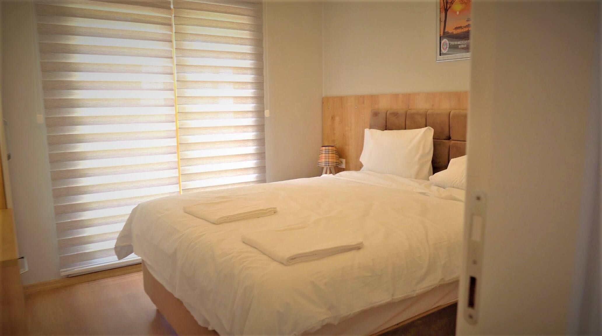 Quarto de hotel com cama, mesa de cabeceira, luminária e janela com persianas.