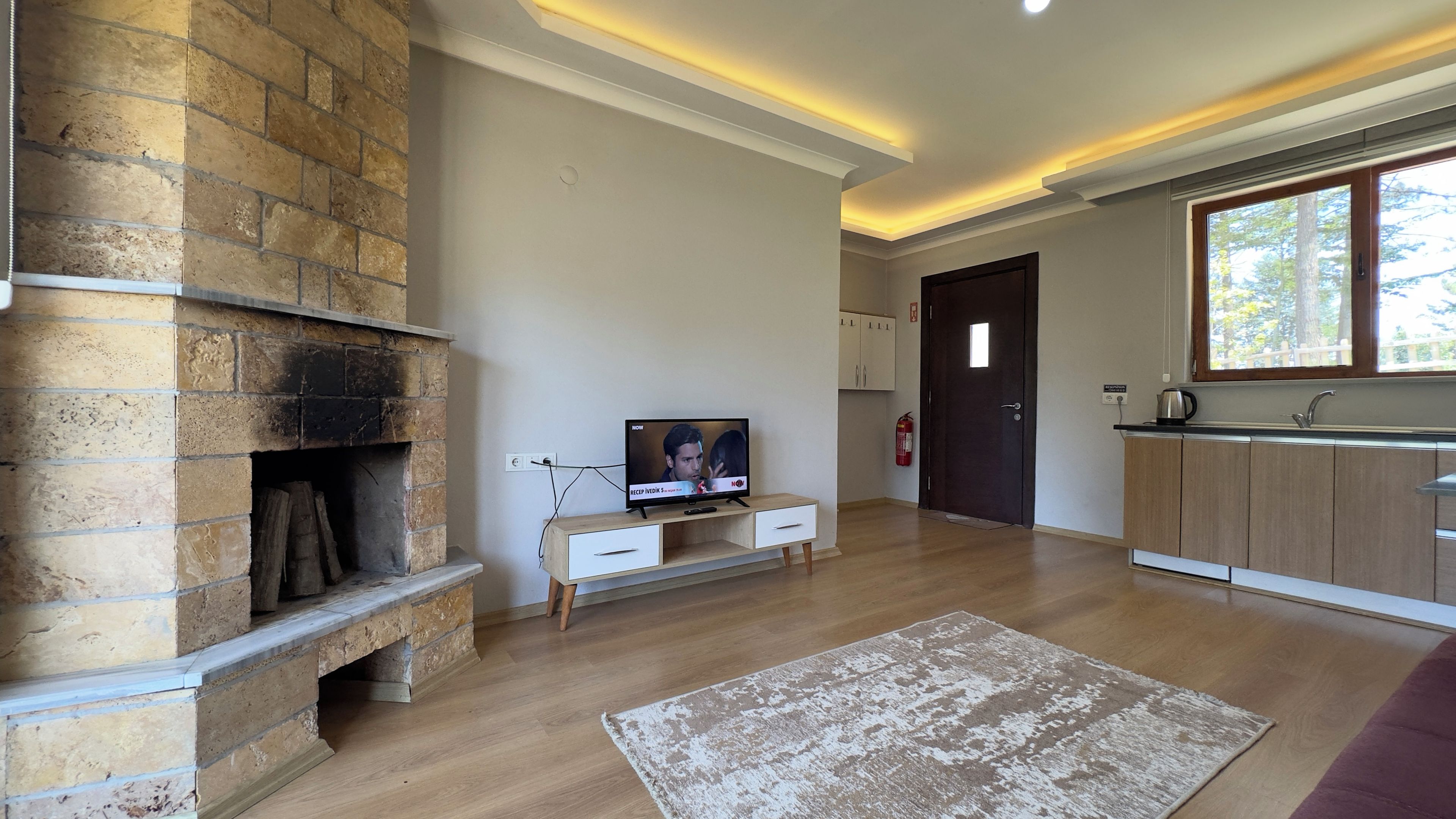 Uma sala de estar com lareira de pedra, televisão, armário de madeira e um espaço de cozinha com janela.