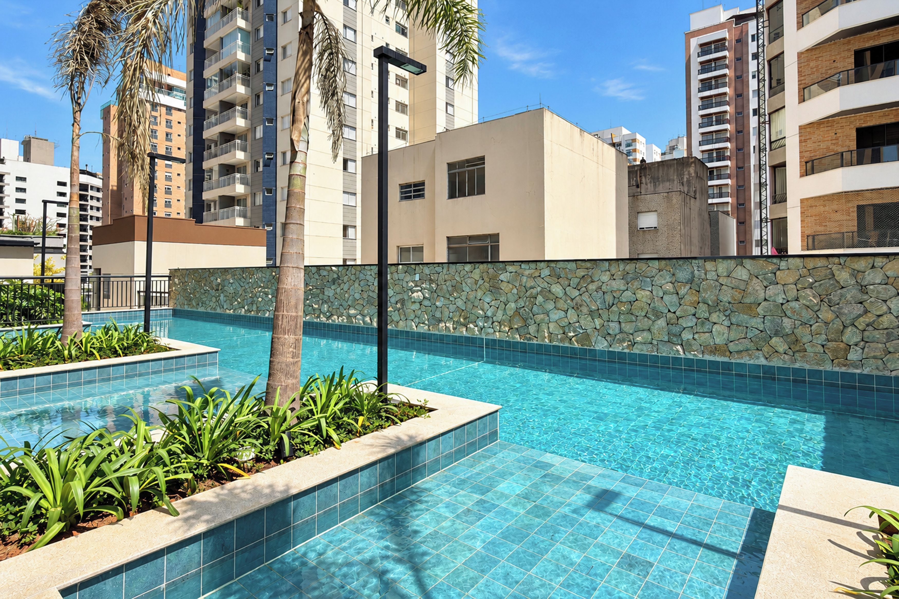 Piscina com água azul cristalina, cercada por um muro de pedra e ladeada por um deck revestido com azulejos.