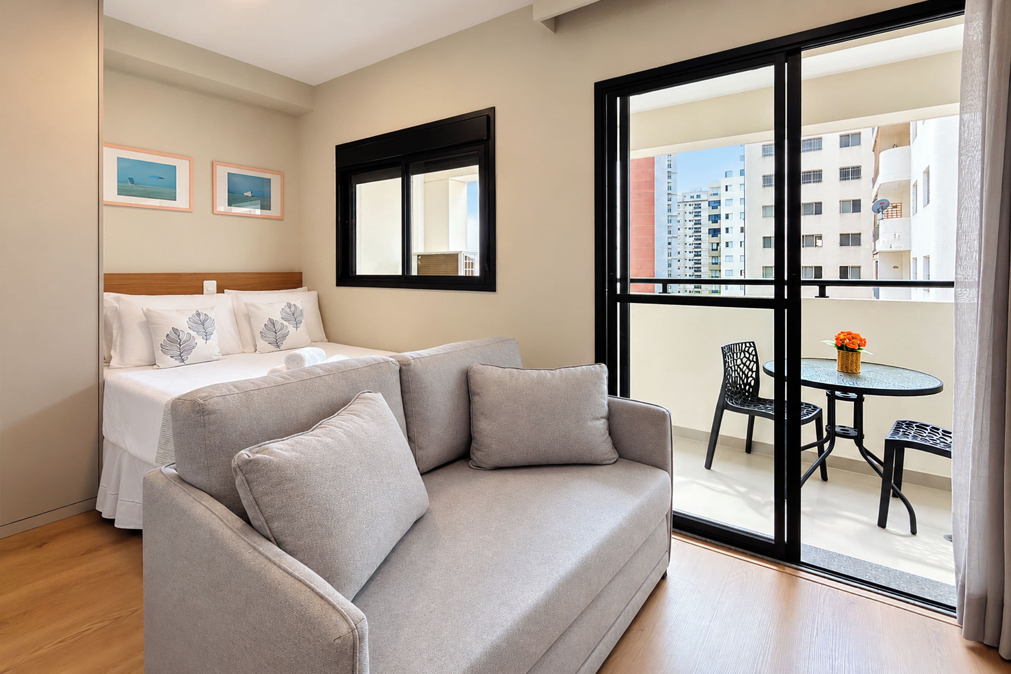 Quarto de hotel com cama, sofá, varanda com mesa e cadeiras, e vista para prédios.