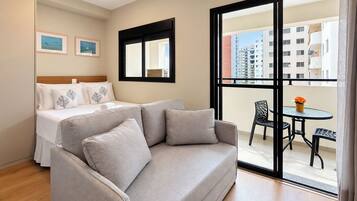 Quarto de hotel com cama, sofá, varanda com mesa e cadeiras, e vista para prédios.