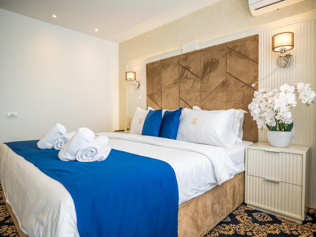 Quarto de hotel com uma cama grande, uma mesa de cabeceira, um abajur fixado na parede e um vaso com flores brancas.