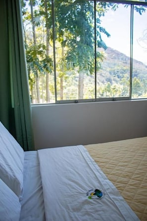 Cama com cobertura acolchoada e travesseiro, janela com vista para árvores e colinas, e cortina parcialmente aberta.