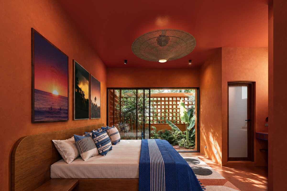 Quarto com cama, almofadas, luminária pendente e vista para o verde através de uma porta.